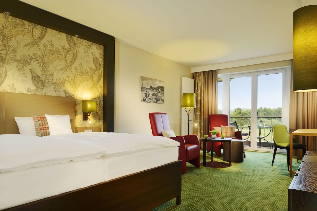 Quarto de hotel com uma cama grande, uma escrivaninha, duas cadeiras, uma pequena mesa com uma planta e uma varanda com vista.