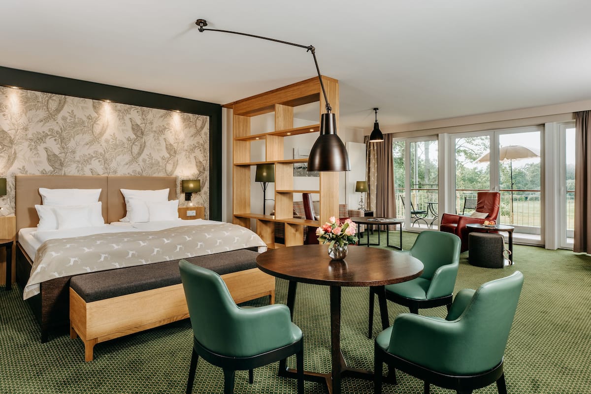Quarto de hotel com cama, mesas de cabeceira, escrivaninha com cadeiras, varanda com mesa e cadeiras, e uma janela grande com vista para árvores.
