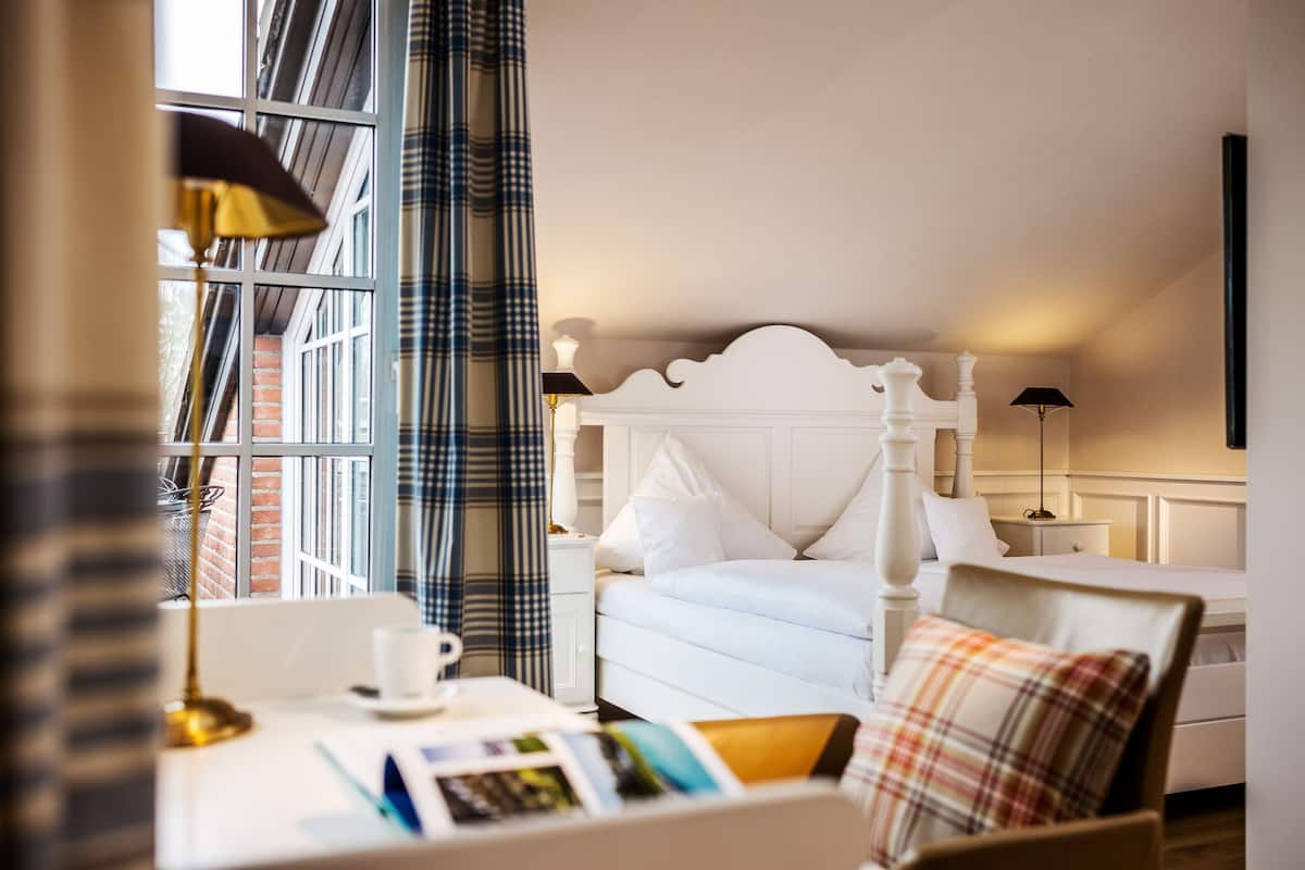 Quarto de hotel com cama branca, mesa de cabeceira, abajur, janela com cortinas e vista para um edifício de tijolos.