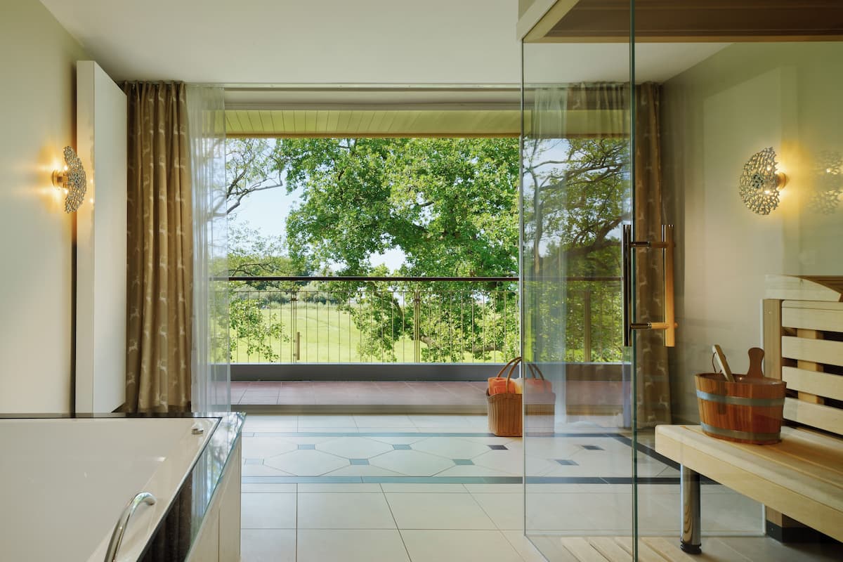 Uma casa de banho moderna com divisória de vidro, um banco de madeira e vista para uma varanda com árvores.