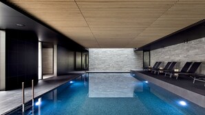 Uma piscina interior moderna com teto de madeira, paredes de pedra e espreguiçadeiras ao longo da borda.