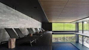 Área de piscina interior moderna com espreguiçadeiras alinhadas contra uma parede, teto de madeira e vista para o exterior.