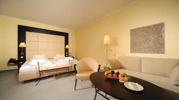 Quarto de hotel com cama, sofá, mesa de centro com frutas e uma obra de arte na parede.