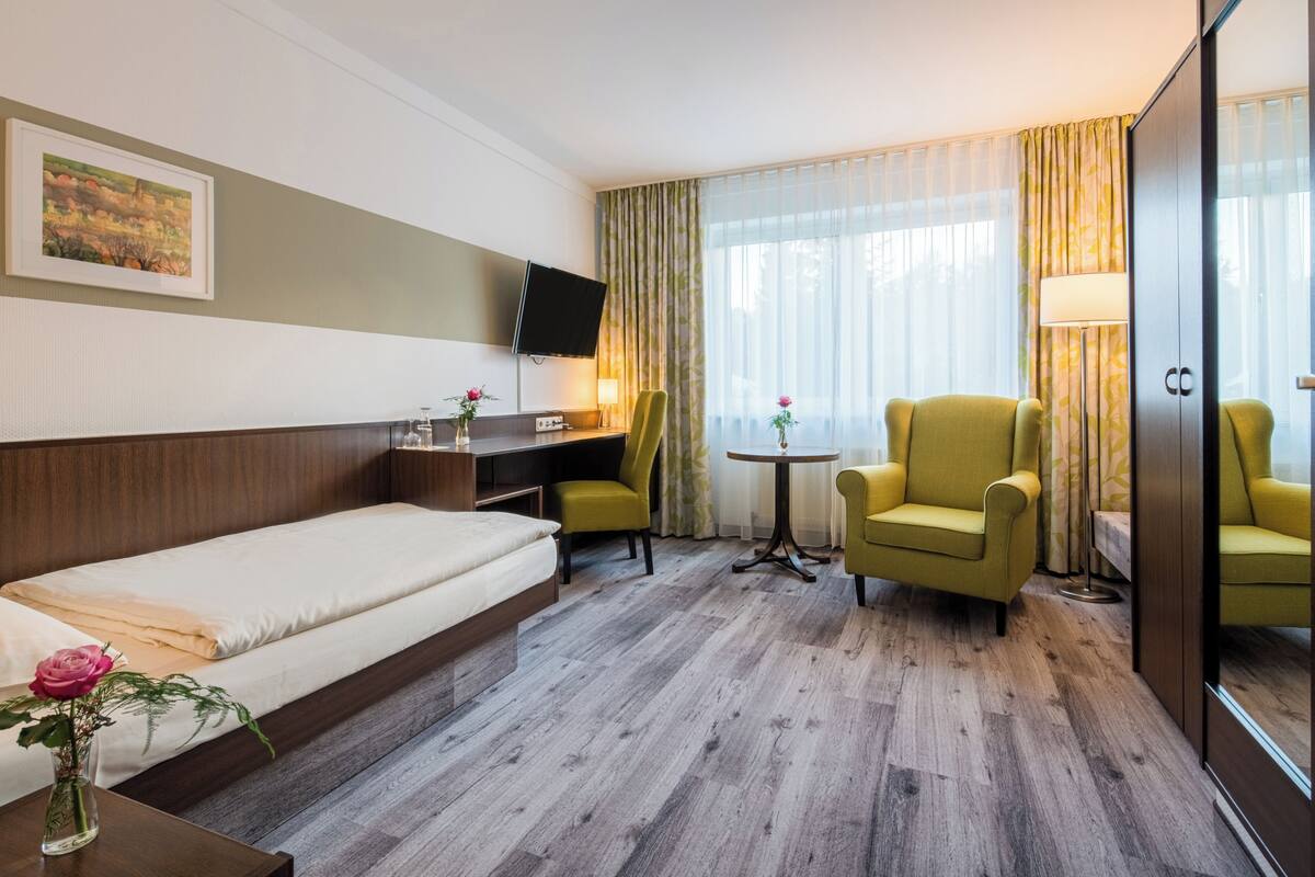 Quarto de hotel com cama, escrivaninha, televisão, duas cadeiras, uma mesinha e um espelho.