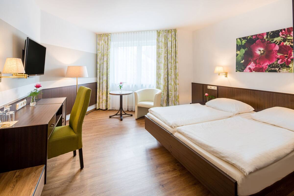 Quarto de hotel com cama, escrivaninha, cadeira, mesinha de centro com um vaso e televisão fixada na parede.