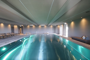 Piscina interior moderna com superfície de água cristalina, cadeiras de espreguiçadeira e área de estar com almofadas.