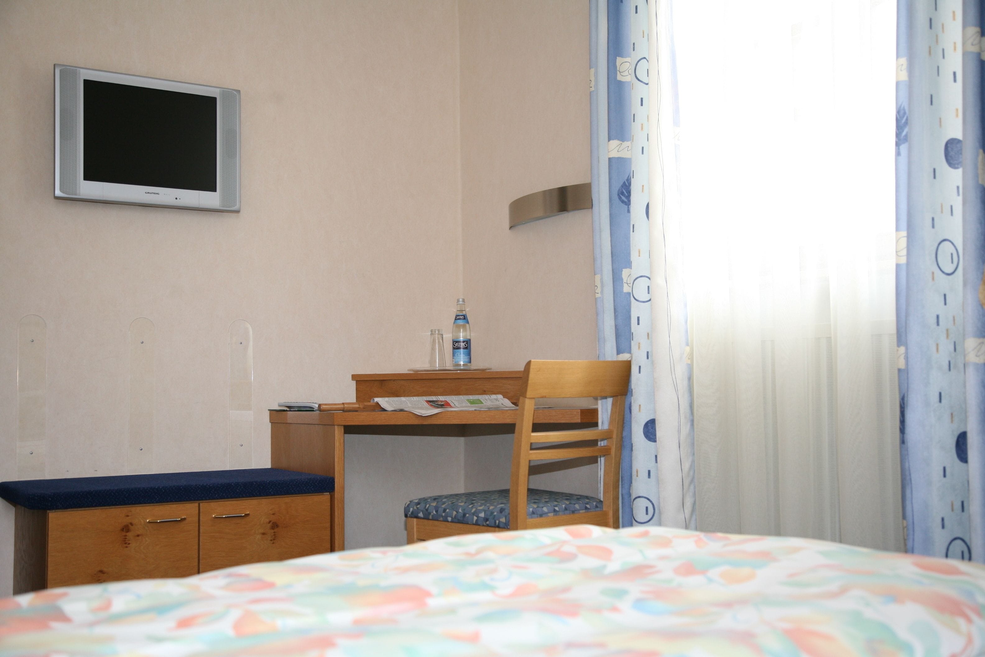 Quarto de hotel com uma cama, uma escrivaninha com cadeira, televisão e janela com cortinas.