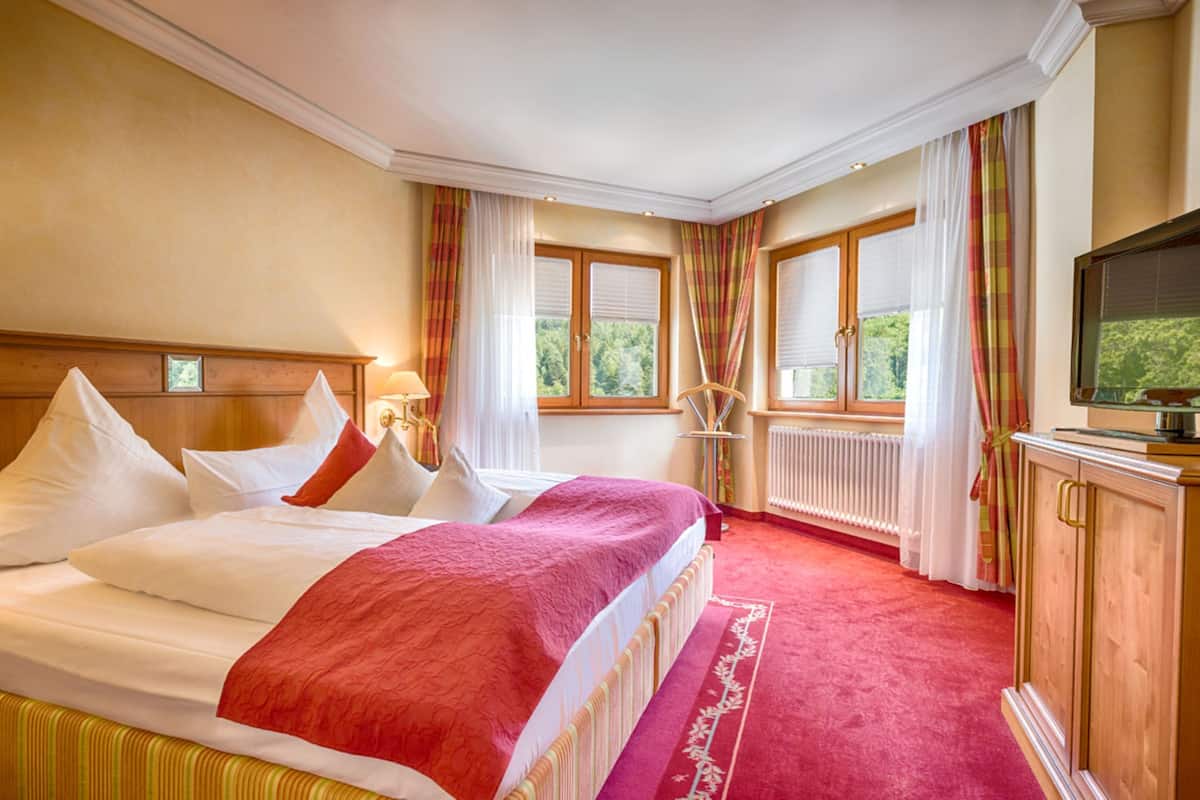 Quarto de hotel com uma cama grande, cabeceira de madeira, televisão e vista para o verde lá fora.