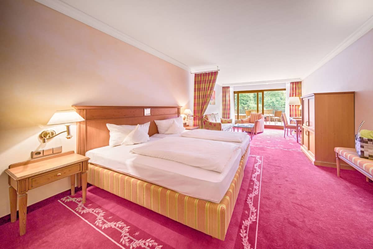 Quarto de hotel com uma cama grande, uma mesinha de cabeceira de madeira, uma escrivaninha e uma cadeira. Há uma janela com cortinas e vista para o exterior.