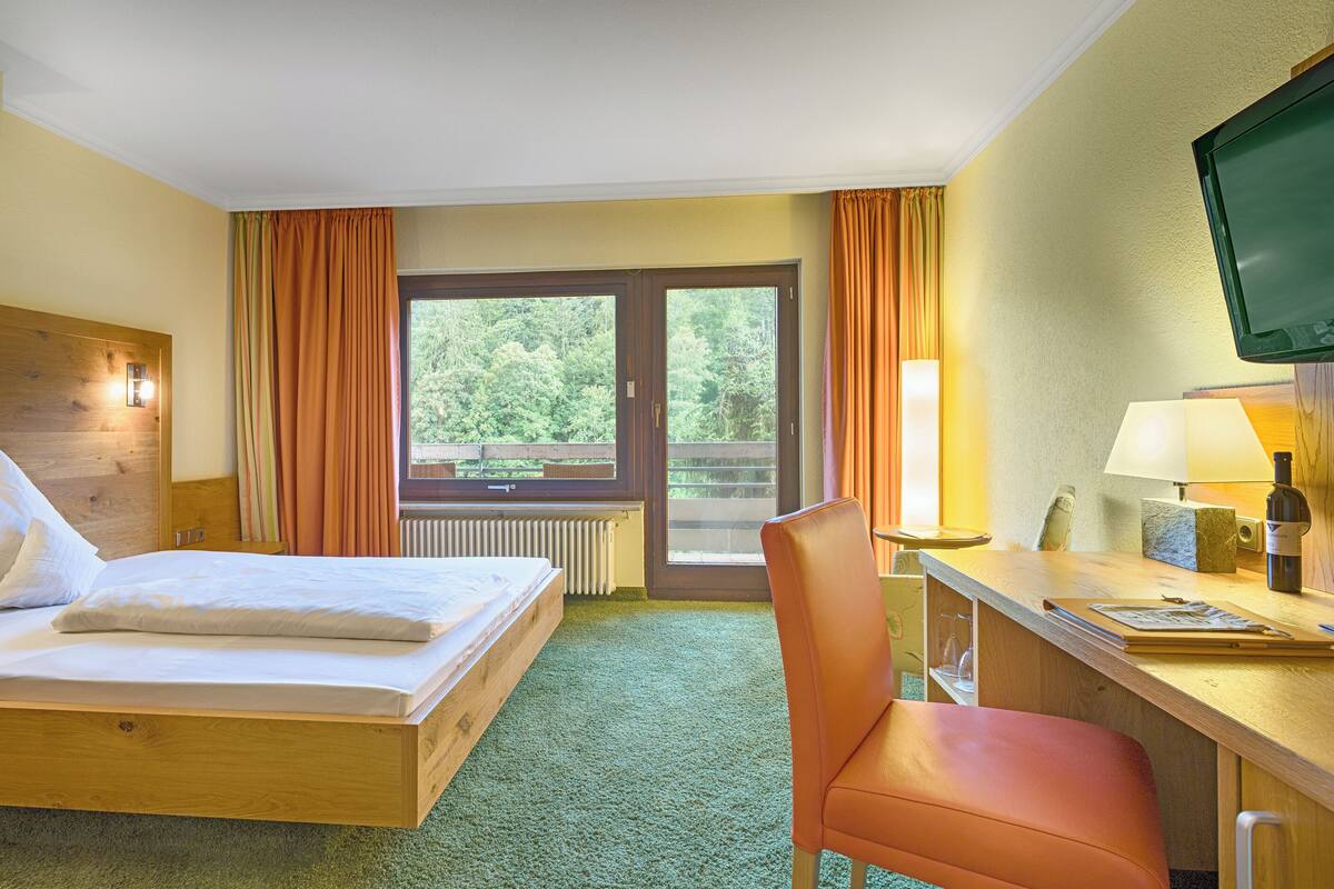 Quarto de hotel com cama, escrivaninha, cadeira, televisão e janela com cortinas.