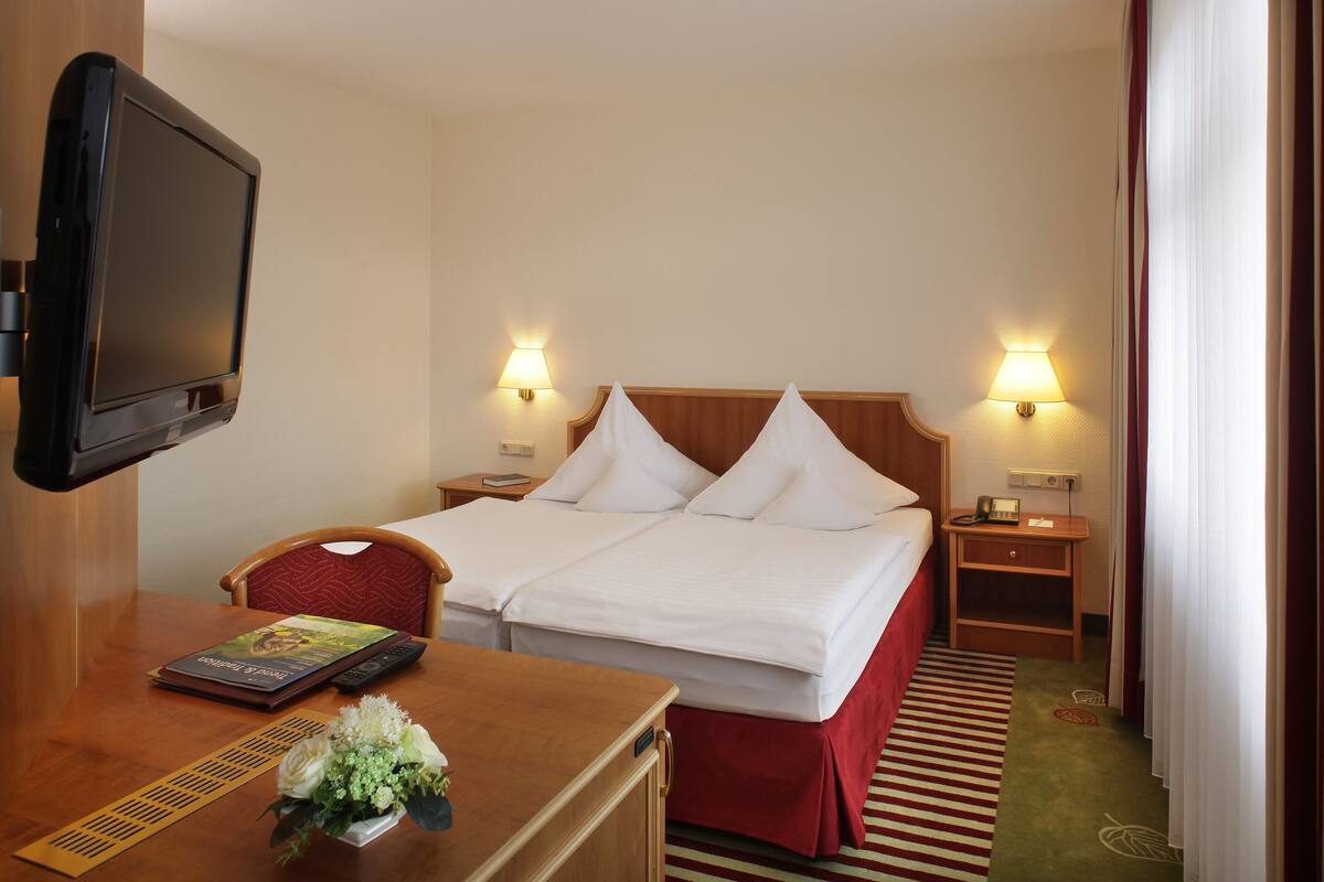 Quarto de hotel com cama, mesas de cabeceira, escrivaninha com luminária, televisão e um vaso de flores.