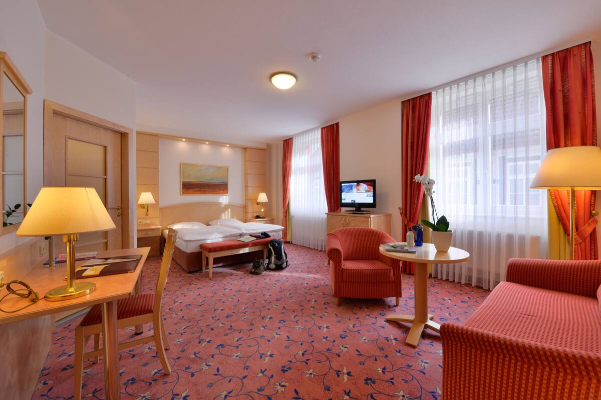 Quarto de hotel com cama, escrivaninha com luminária, cadeira, mesinha com planta e televisão.