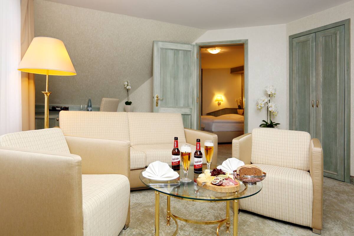 Quarto de hotel com sofá bege, mesa de centro de vidro com comida e bebidas, e uma luminária.