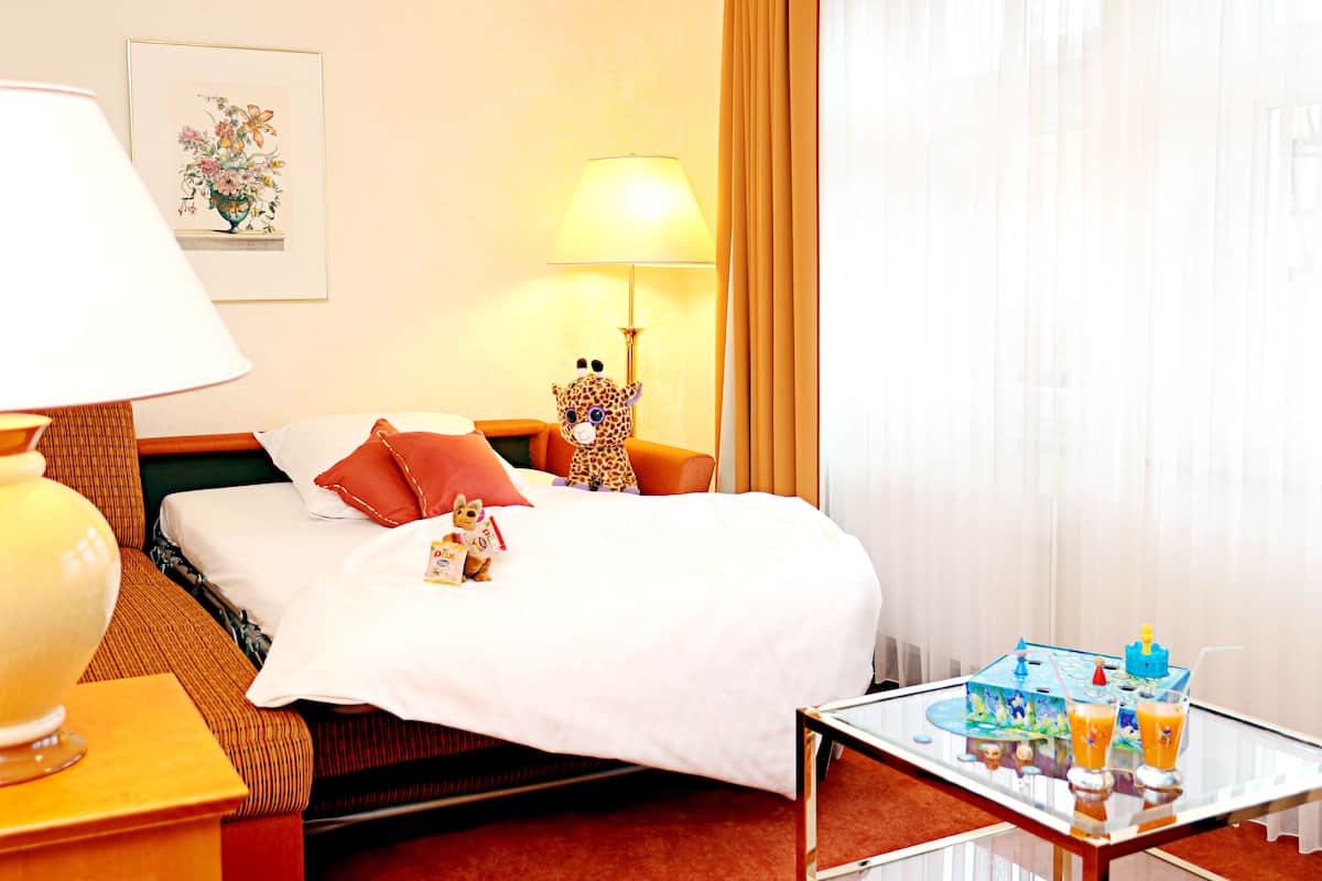 Quarto de hotel com uma cama, mesa de cabeceira com abajur, mesa de vidro com um jogo de tabuleiro e bebidas, e um bichinho de pelúcia de girafa na cama.