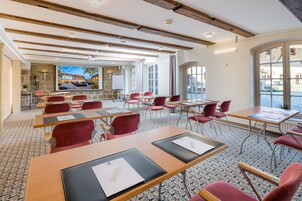 Sala de conferências com mesas, cadeiras, tela de projeção e parede de pedra com vista para edifícios.
