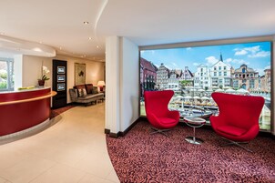 Um saguão de hotel com balcão de recepção, área de estar com cadeiras vermelhas, carpete estampado e uma grande janela com vista para edifícios históricos.