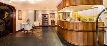 Uma área de recepção com balcão curvo de madeira, espaço de estar com duas cadeiras, uma pequena mesa e um vaso de planta.