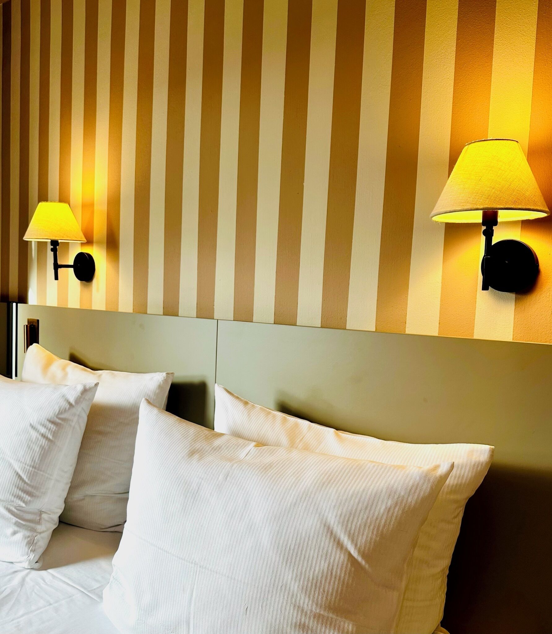 Quarto de hotel com parede listrada, duas luminárias fixadas na parede e uma cama com almofadas brancas.