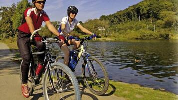 Duas pessoas andando de bicicleta em um caminho pavimentado ao lado de um corpo d'água, com árvores e uma colina ao fundo.