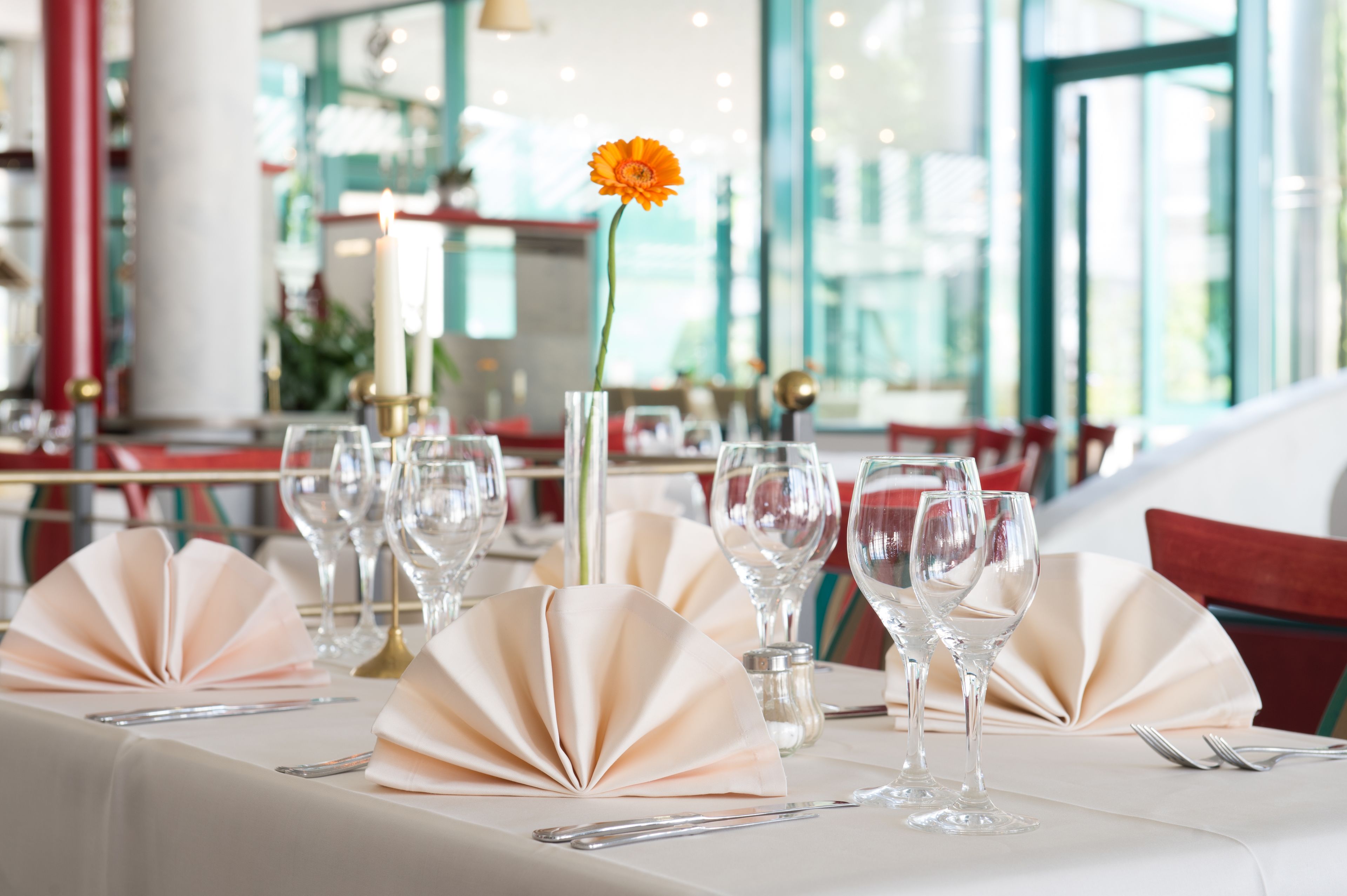 Mesa de jantar posta com toalhas brancas, copos de vidro elegantes e um arranjo central com uma única flor laranja.