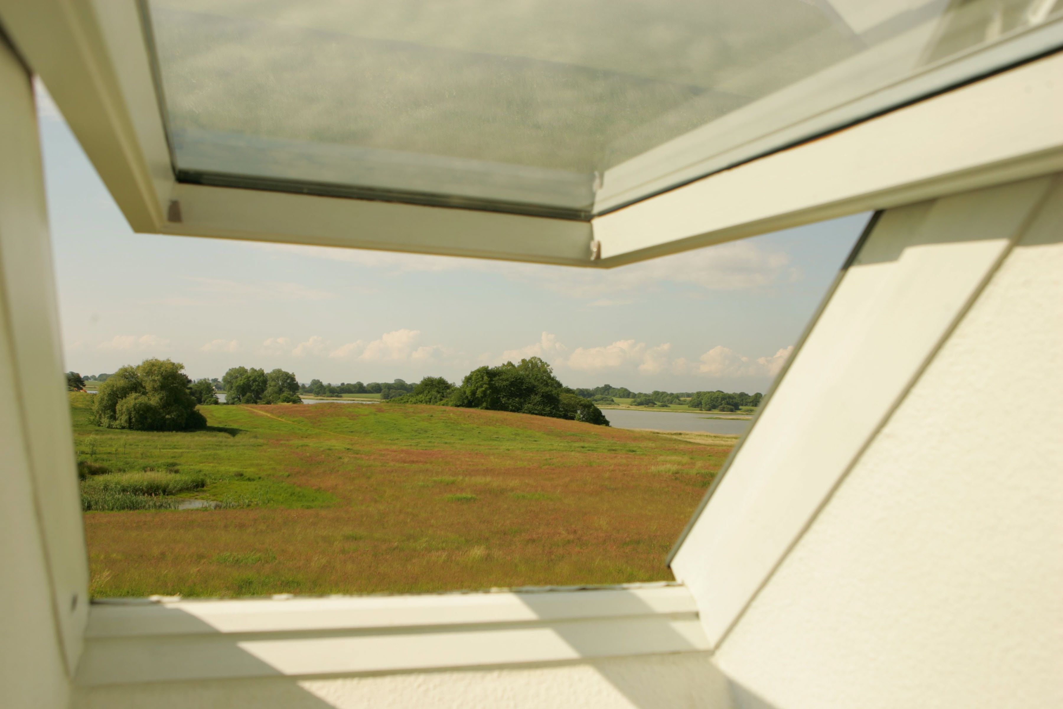 Visão através de uma janela com moldura branca, com vista para um campo gramado e um corpo d'água.