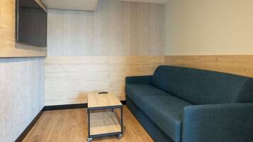Quarto com piso de madeira, sofá azul, pequena mesa de madeira com controle remoto e ar-condicionado fixado na parede.