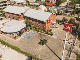 Um edifício de vários andares com um grande letreiro vermelho 'ibis', um estacionamento com vagas demarcadas e uma grande estrutura circular à esquerda.