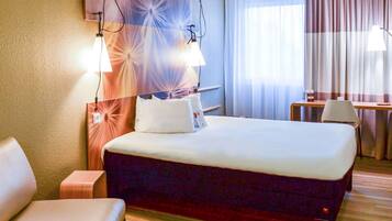 Quarto de hotel com cama, mesa de cabeceira, cadeira, escrivaninha, luminária e cortinas.