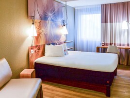 Quarto de hotel com cama, mesa de cabeceira, cadeira, escrivaninha, luminária e cortinas.