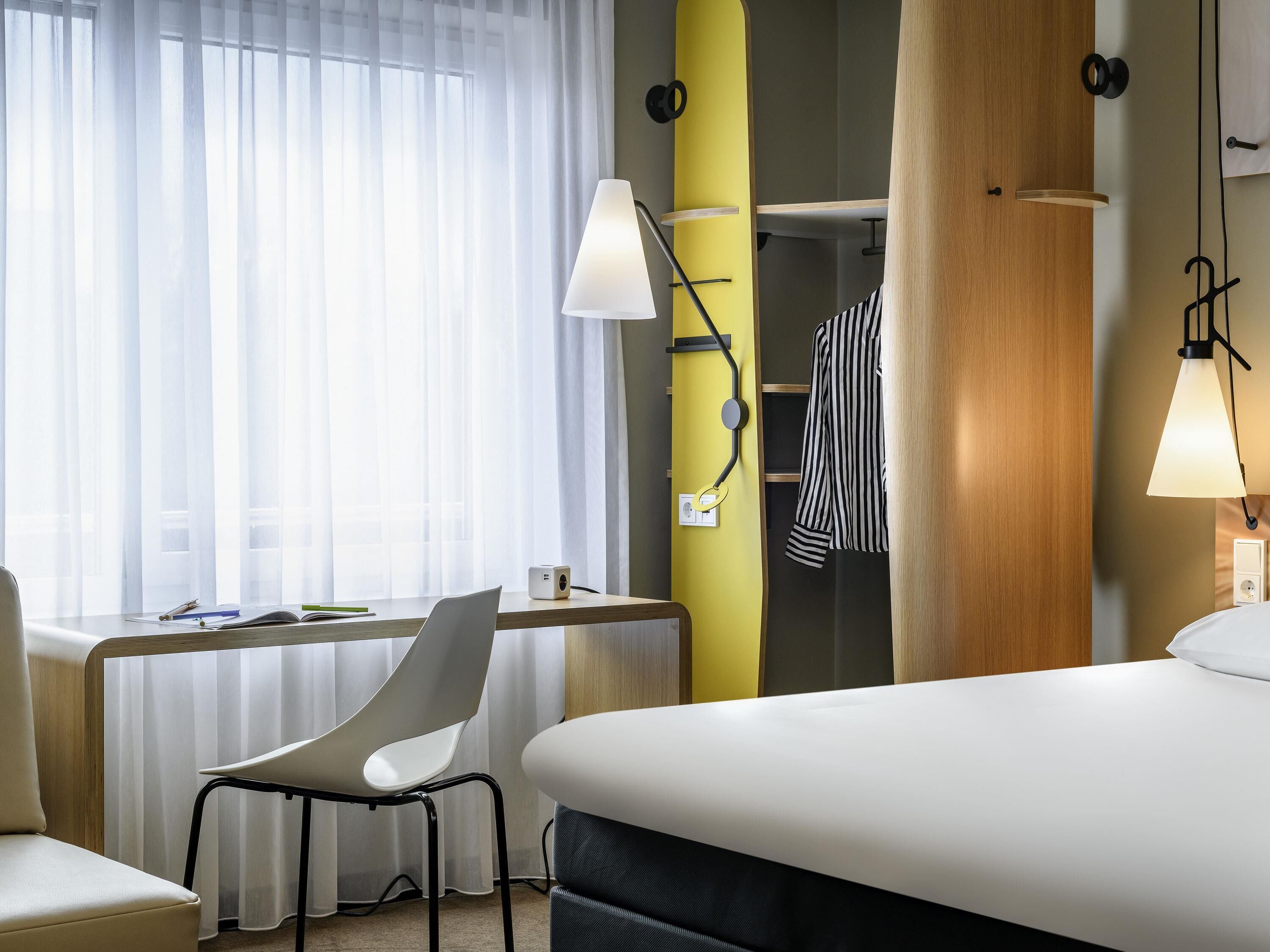 Quarto de hotel com cama, escrivaninha com cadeira, luminária, guarda-roupa e janela com cortinas finas.