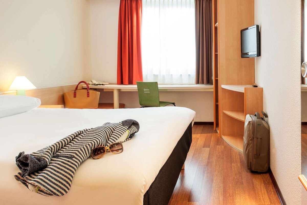 Quarto de hotel com cama, escrivaninha, cadeira, televisão e mala.