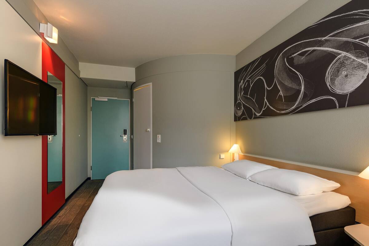 Quarto de hotel com uma cama grande, televisão na parede, um quadro e uma mesa com abajur.
