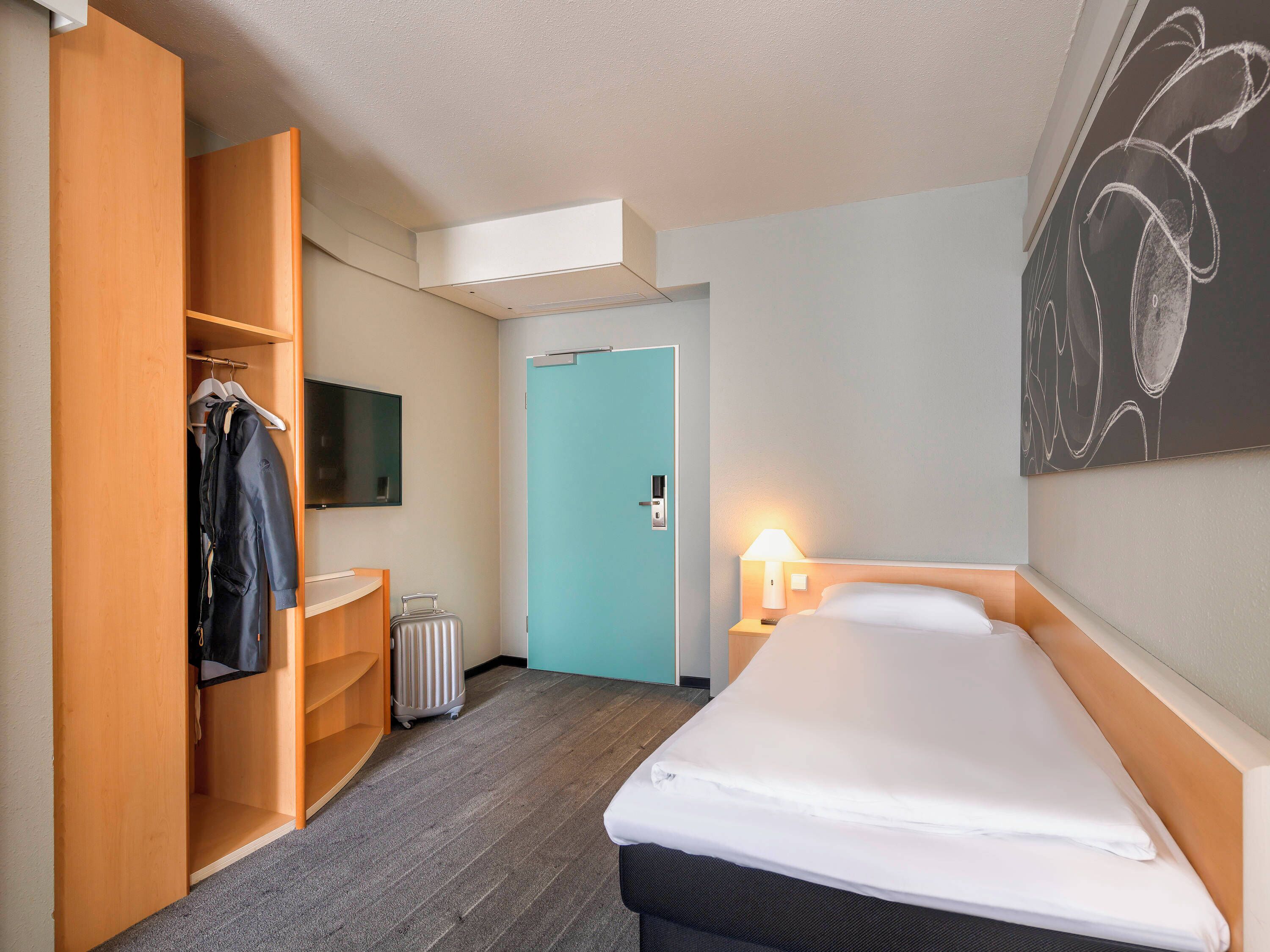 Quarto de hotel com uma cama, um guarda-roupa, uma televisão, uma porta e uma obra de arte na parede.
