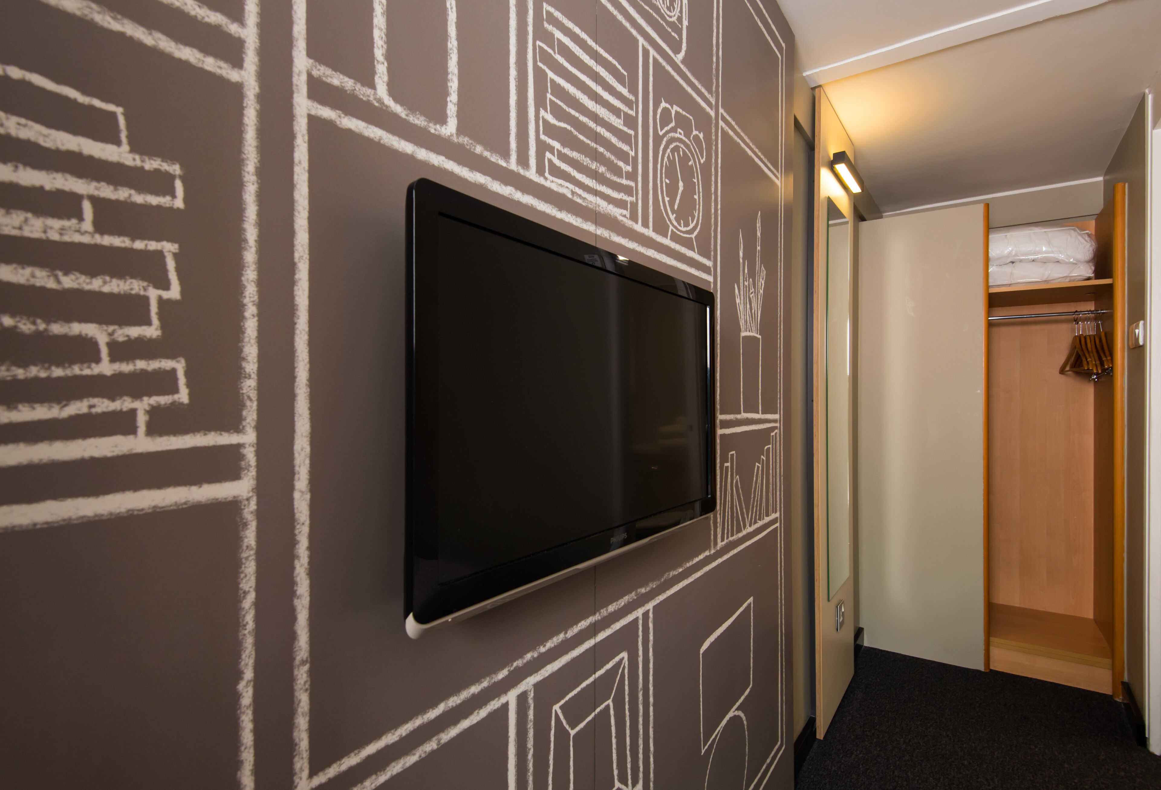 Quarto com televisão de tela plana na parede, guarda-roupa de madeira e piso coberto por carpete.