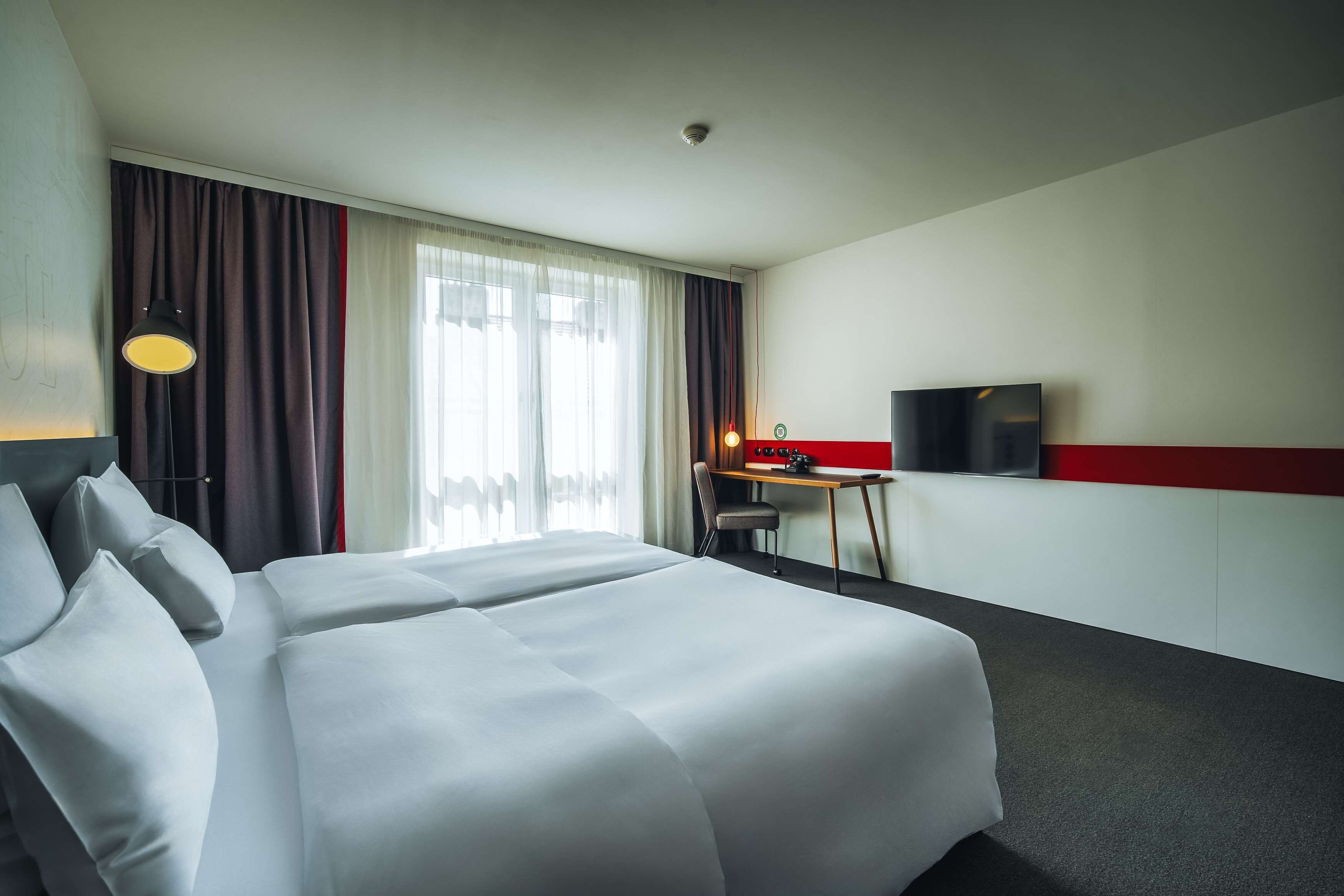 Quarto de hotel com duas camas, uma mesa com cadeira, televisão e janela com cortinas.