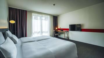 Quarto de hotel com duas camas, uma mesa com cadeira, televisão e janela com cortinas.