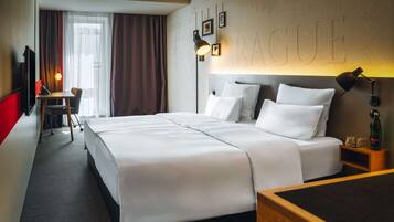 Quarto de hotel com uma cama grande, uma escrivaninha com cadeira, televisão e uma parede com a palavra 'RAGUE' parcialmente visível.