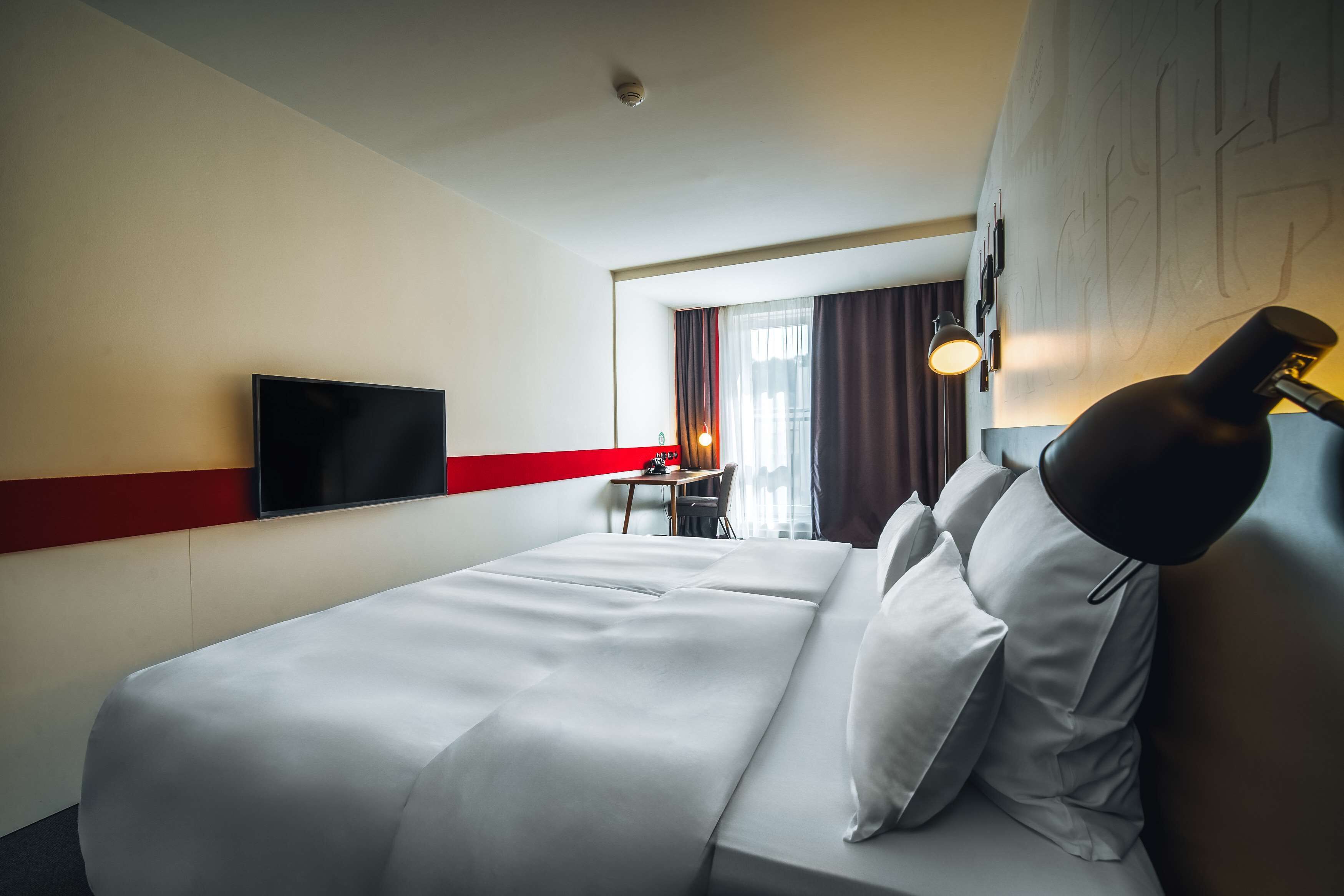 Quarto de hotel com cama, escrivaninha, cadeira, televisão e abajur.