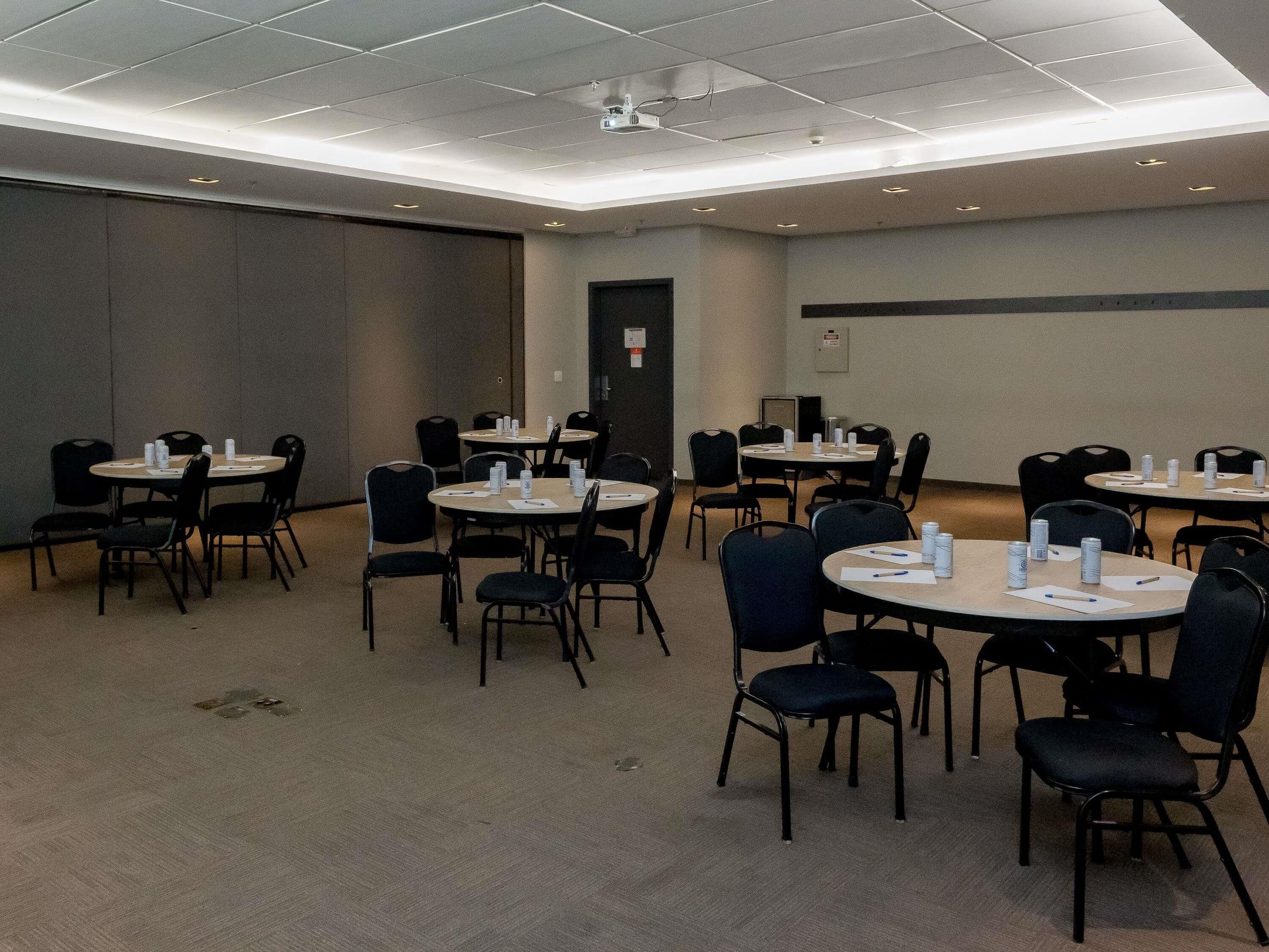 Sala de conferência com várias mesas e cadeiras, equipada com garrafas de água e blocos de notas.