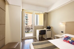 Quarto de hotel com cama, escrivaninha, cadeira e vista da cidade através de uma grande janela.