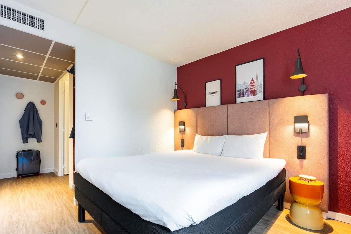 Quarto de hotel com cama, parede em destaque vermelha e uma pequena mesa com abajur.