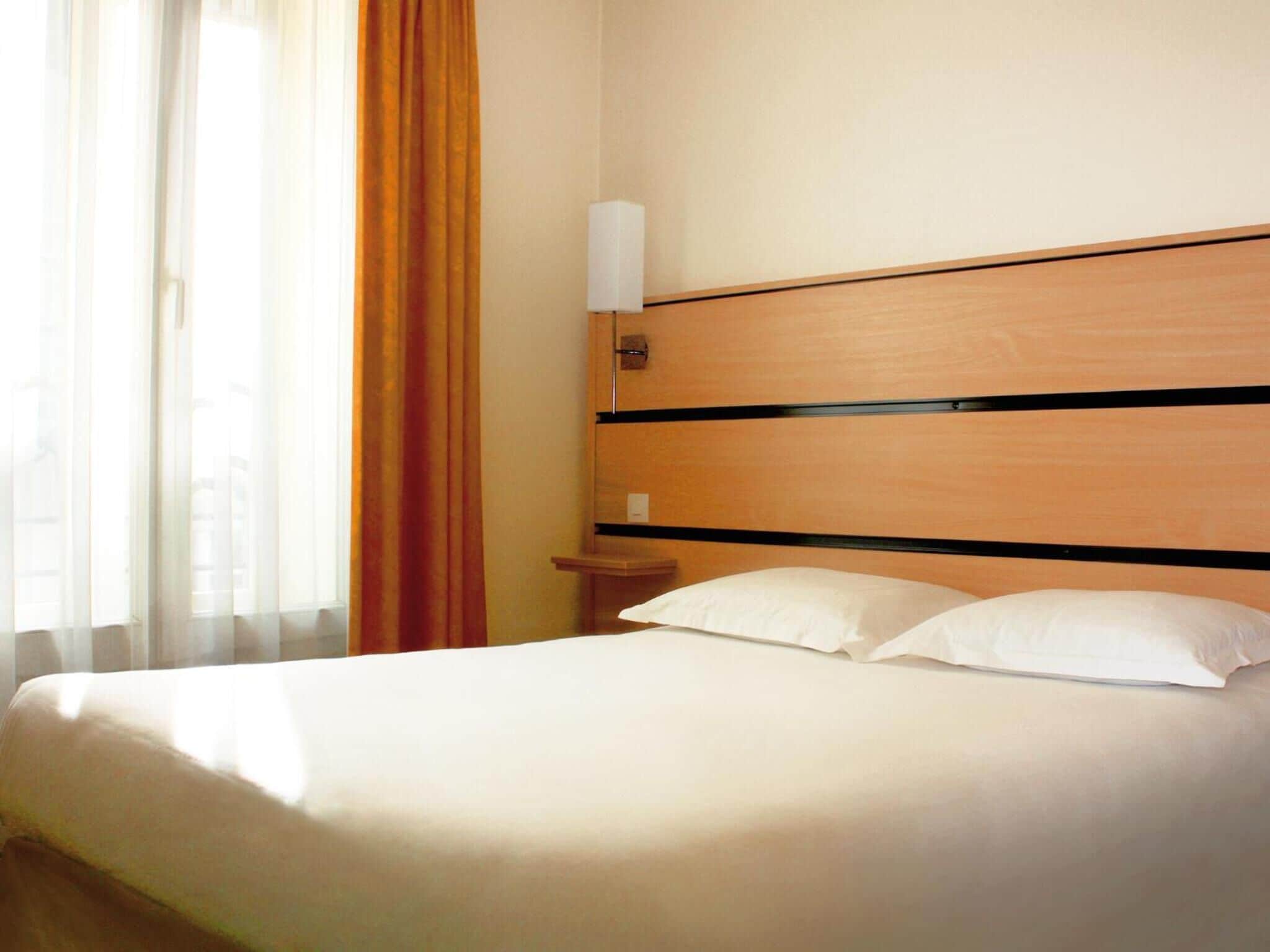 Quarto de hotel com cabeceira de madeira, mesa de cabeceira com abajur e uma cama grande com roupa de cama branca.