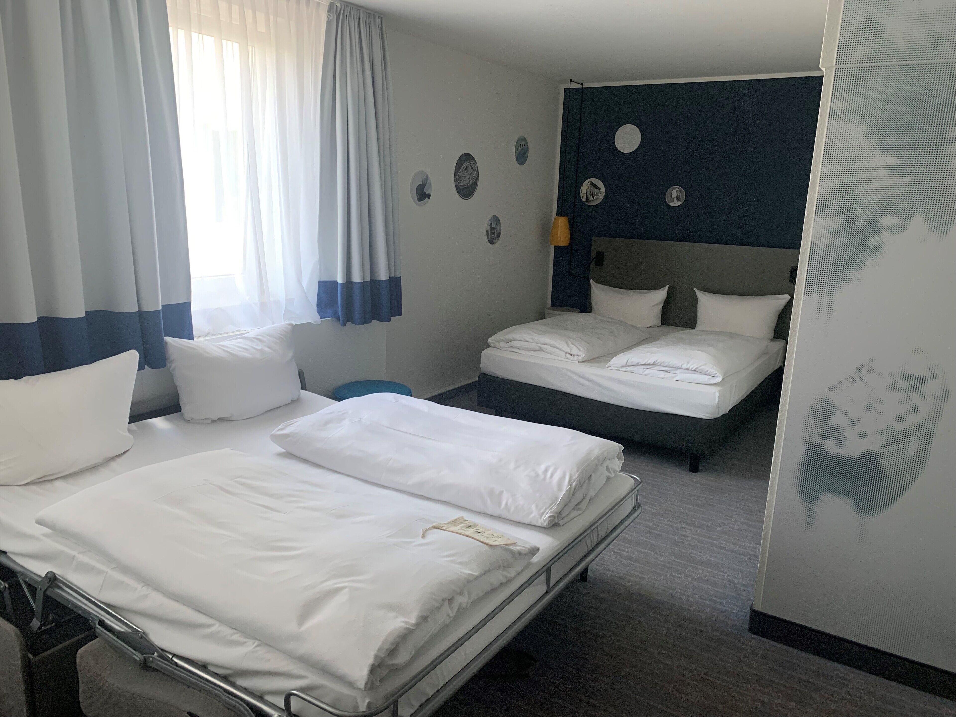 Quarto de hotel com uma cama de casal, uma cama individual, uma mesinha de cabeceira, uma luminária e uma parede com decorações circulares.