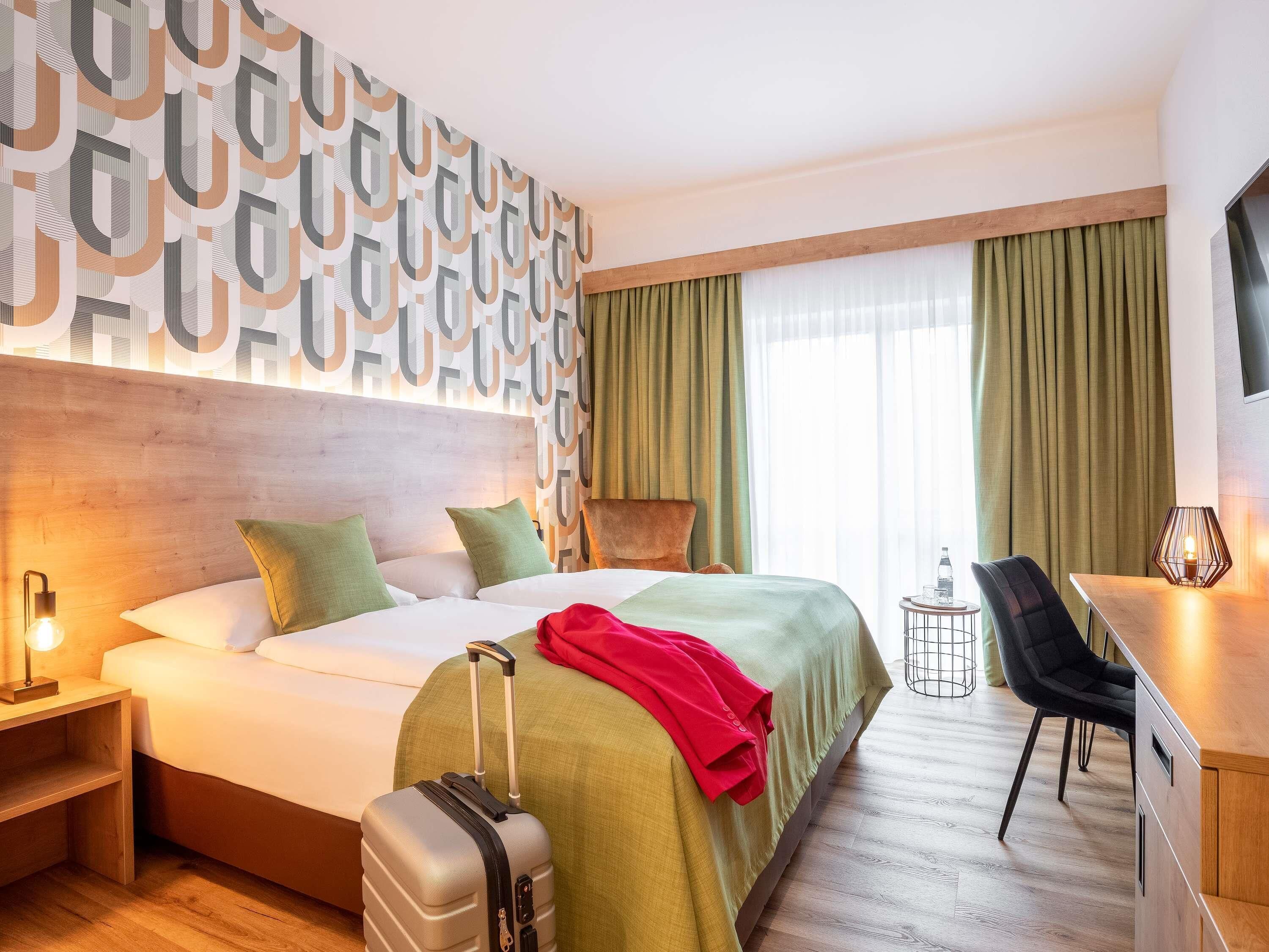 Quarto de hotel com cama, escrivaninha, cadeira, mala e televisão na parede.