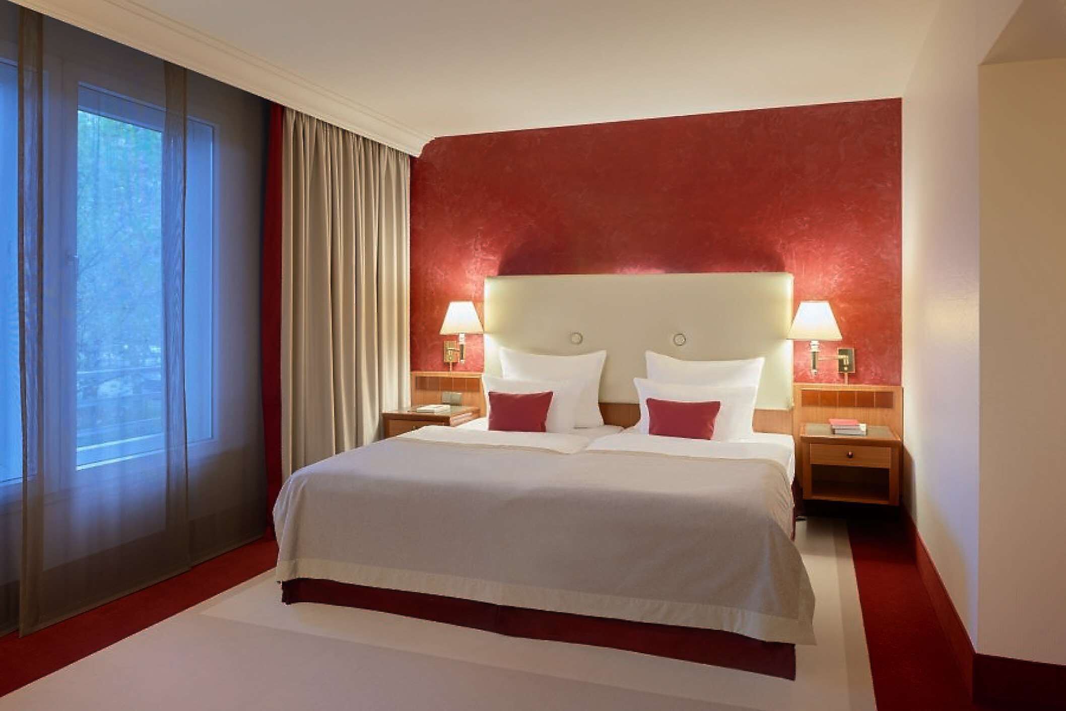 Quarto de hotel com cama grande, duas mesas de cabeceira, um criado-mudo, janela com cortinas e uma parede em vermelho.