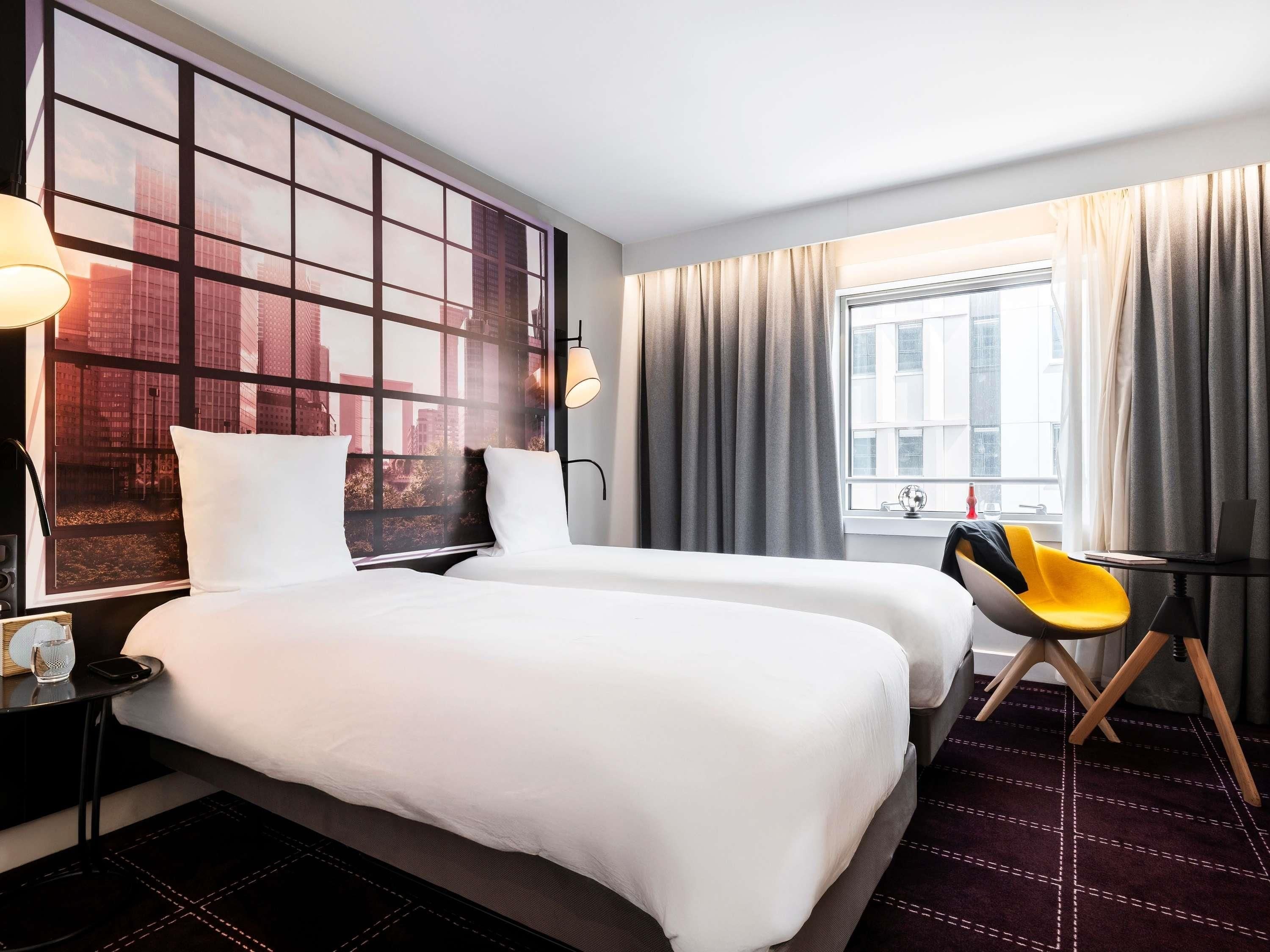 Quarto de hotel com duas camas, uma escrivaninha com cadeira, janela com cortinas e vista para a cidade.