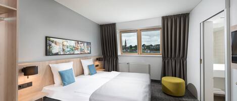 Quarto de hotel com cama, um quadro, janela com cortinas, um banquinho pequeno e vista para o exterior.