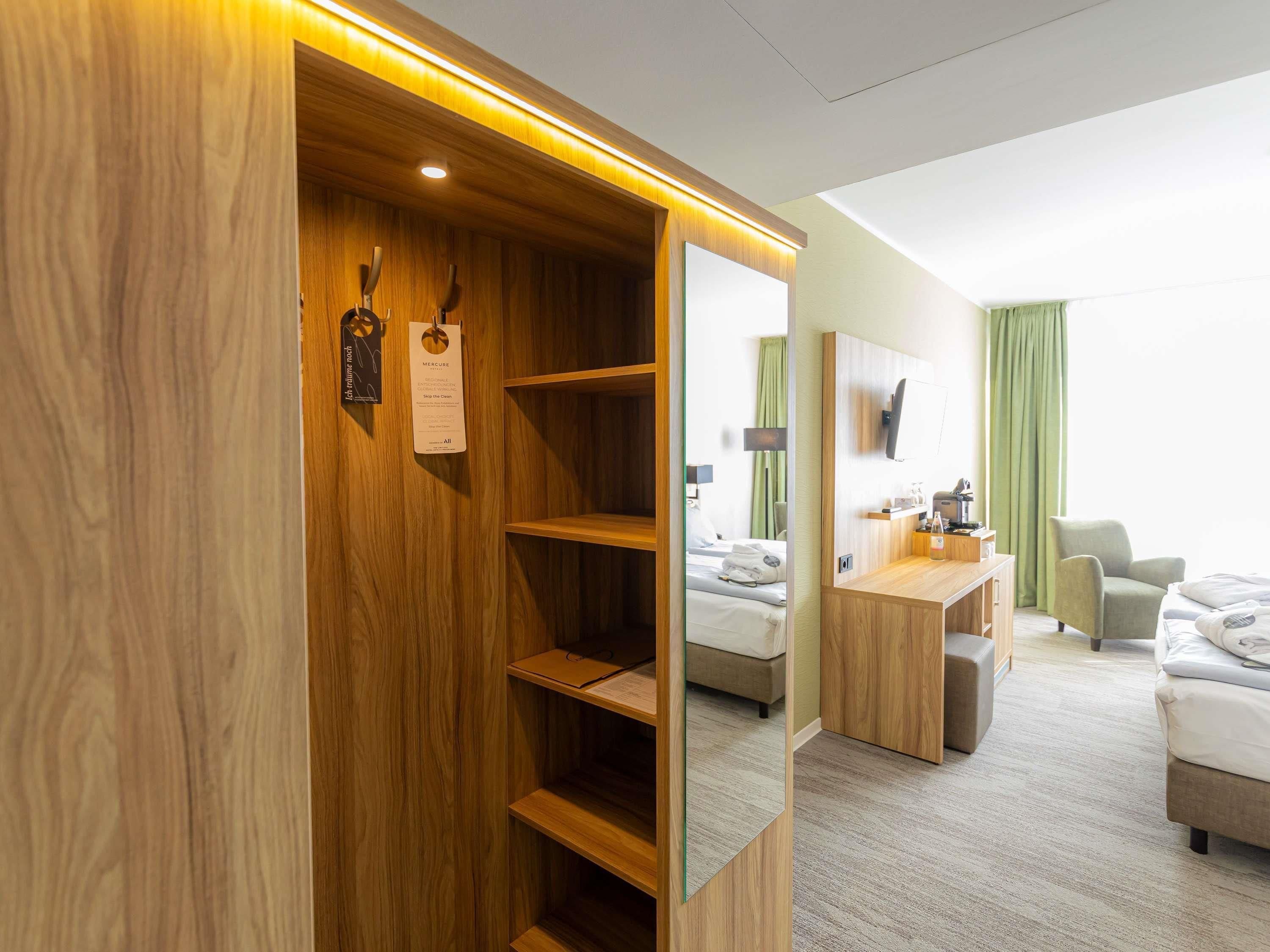 Quarto de hotel com armário de madeira, escrivaninha com cadeira, cama, televisão e espelho.