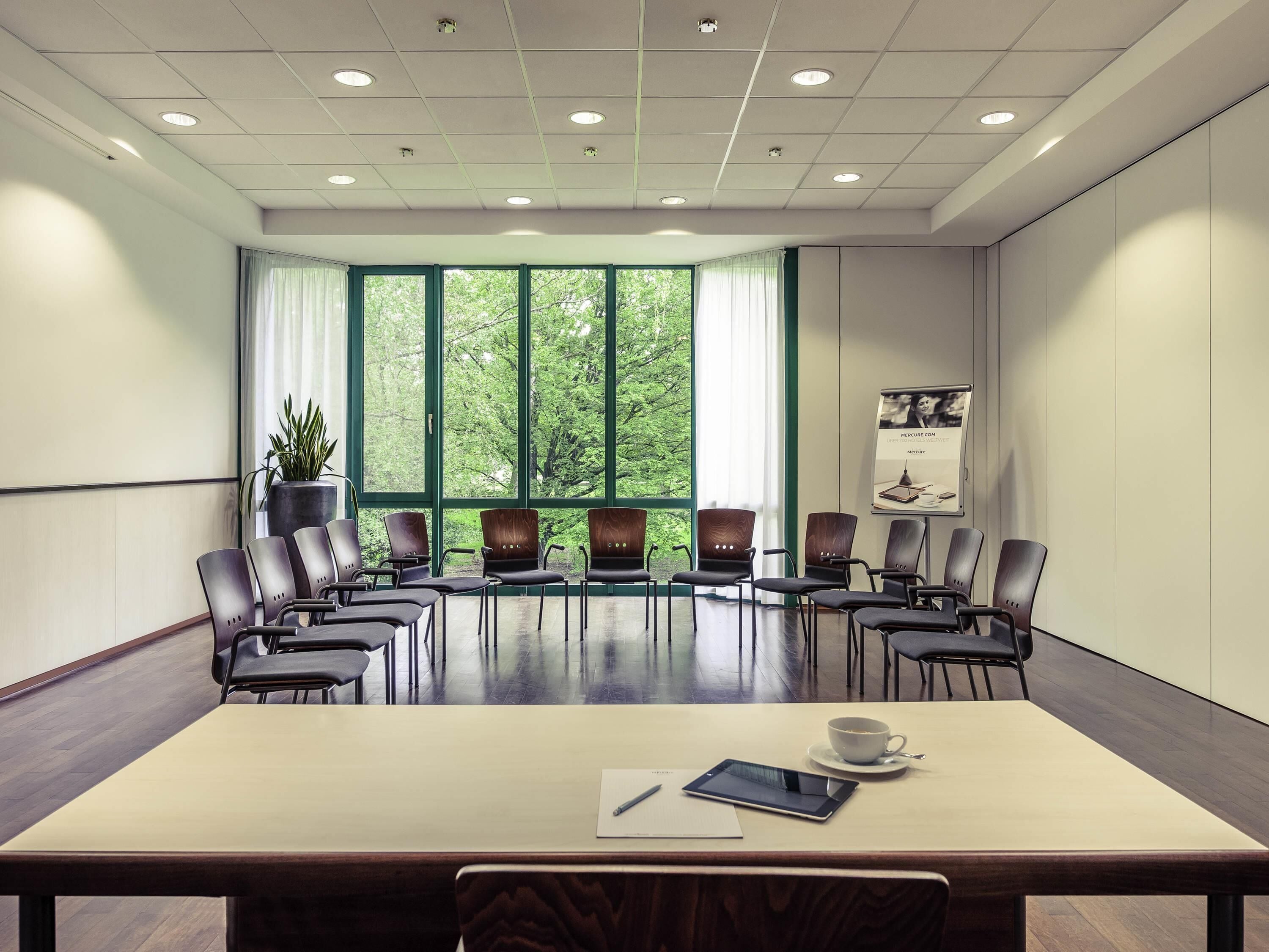 Uma sala de conferências com uma mesa comprida, cadeiras, um vaso de planta e uma janela com vista para árvores.