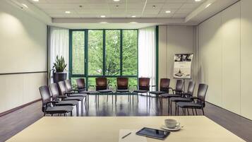 Uma sala de conferências com uma mesa comprida, cadeiras, um vaso de planta e uma janela com vista para árvores.
